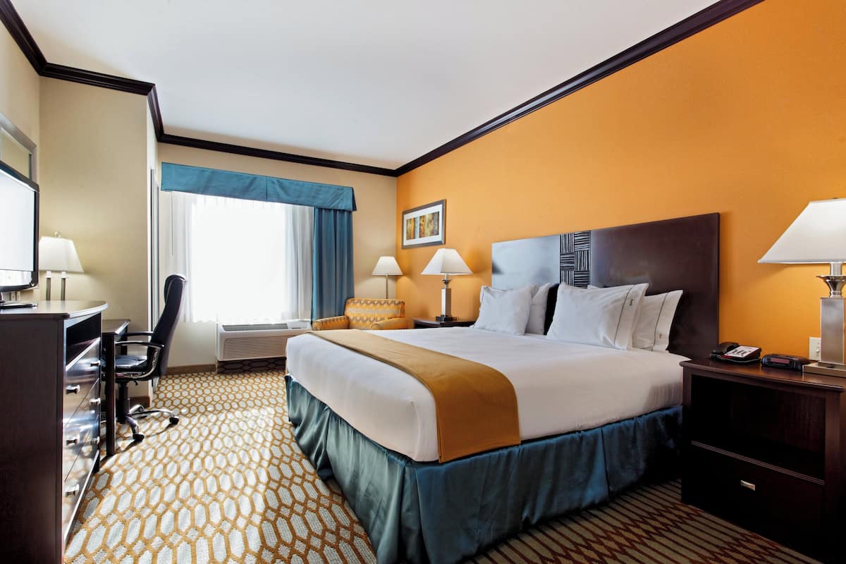 Quarto de hotel com uma cama grande, uma escrivaninha com cadeira, uma luminária de mesa, uma mesa de cabeceira com abajur e uma janela com cortinas.
