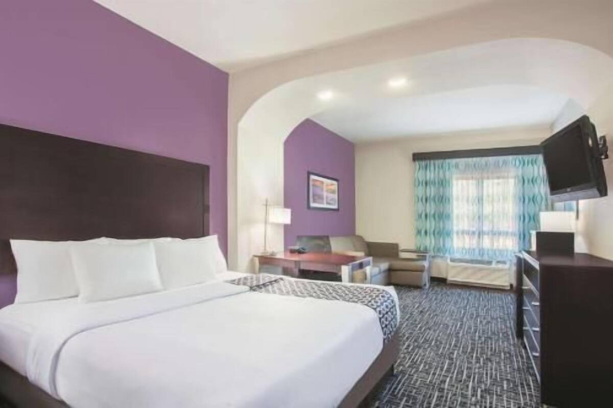 Quarto de hotel com uma cama grande, uma escrivaninha, uma cadeira, uma televisão e uma janela com cortinas.