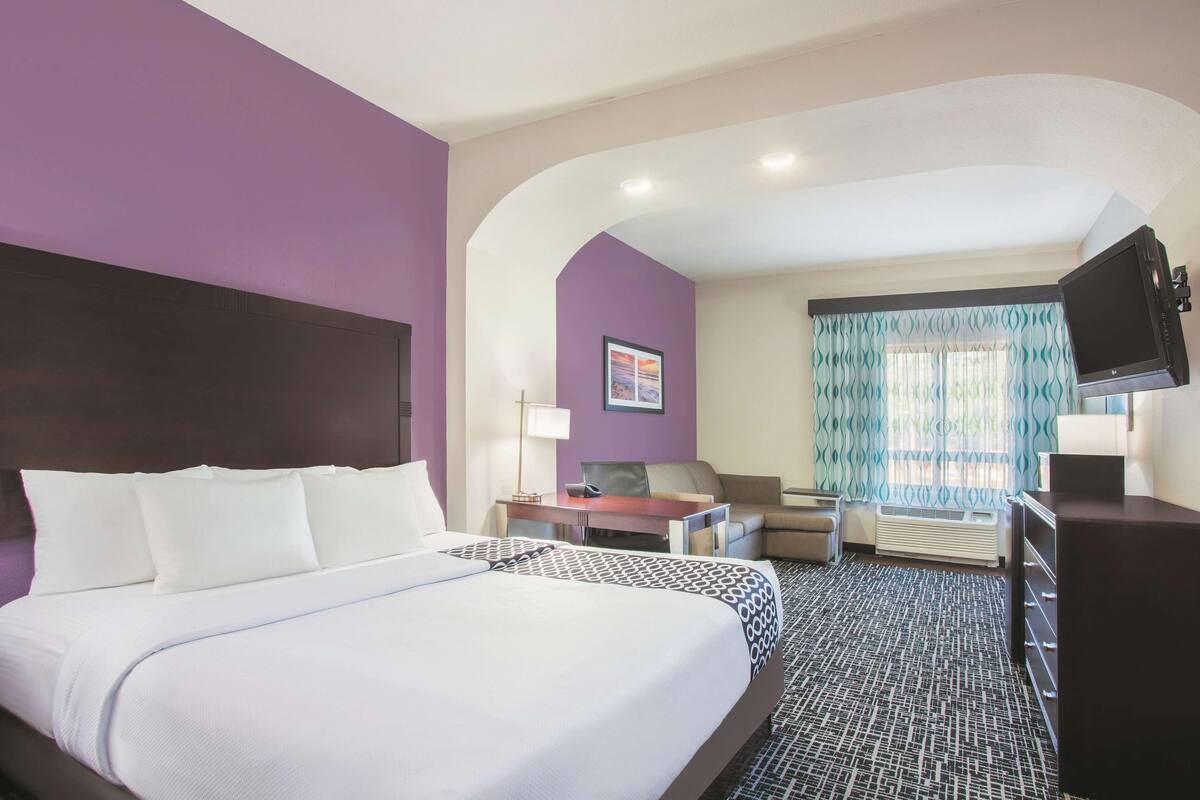 Quarto de hotel com uma cama grande, uma escrivaninha, uma cadeira, uma televisão e uma janela com cortinas.