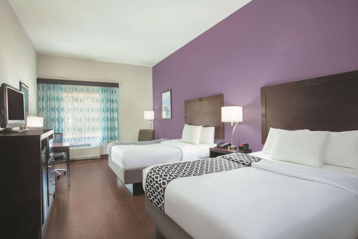 Quarto de hotel com duas camas, uma escrivaninha, uma cadeira, uma televisão e uma janela com cortinas estampadas.