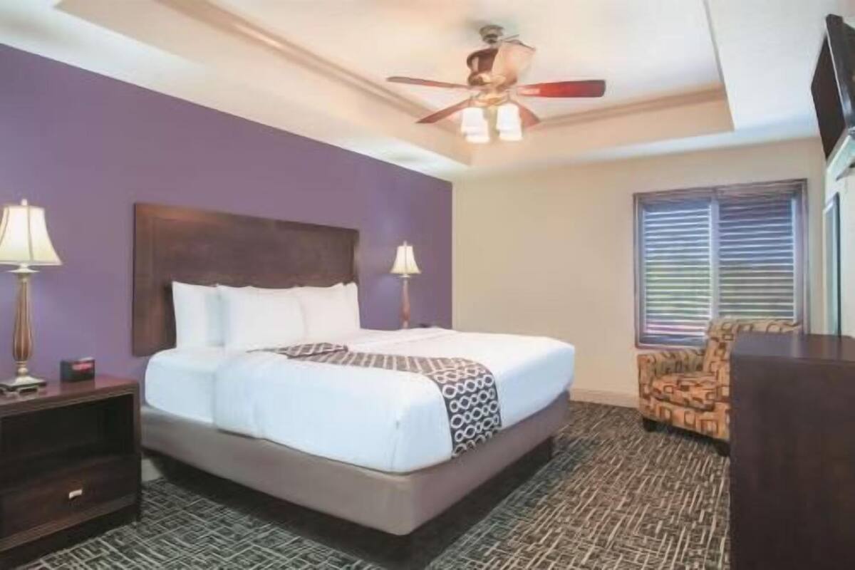 Quarto de hotel com uma cama, duas luminárias de cabeceira, um ventilador de teto, uma janela com persianas e um tapete estampado.