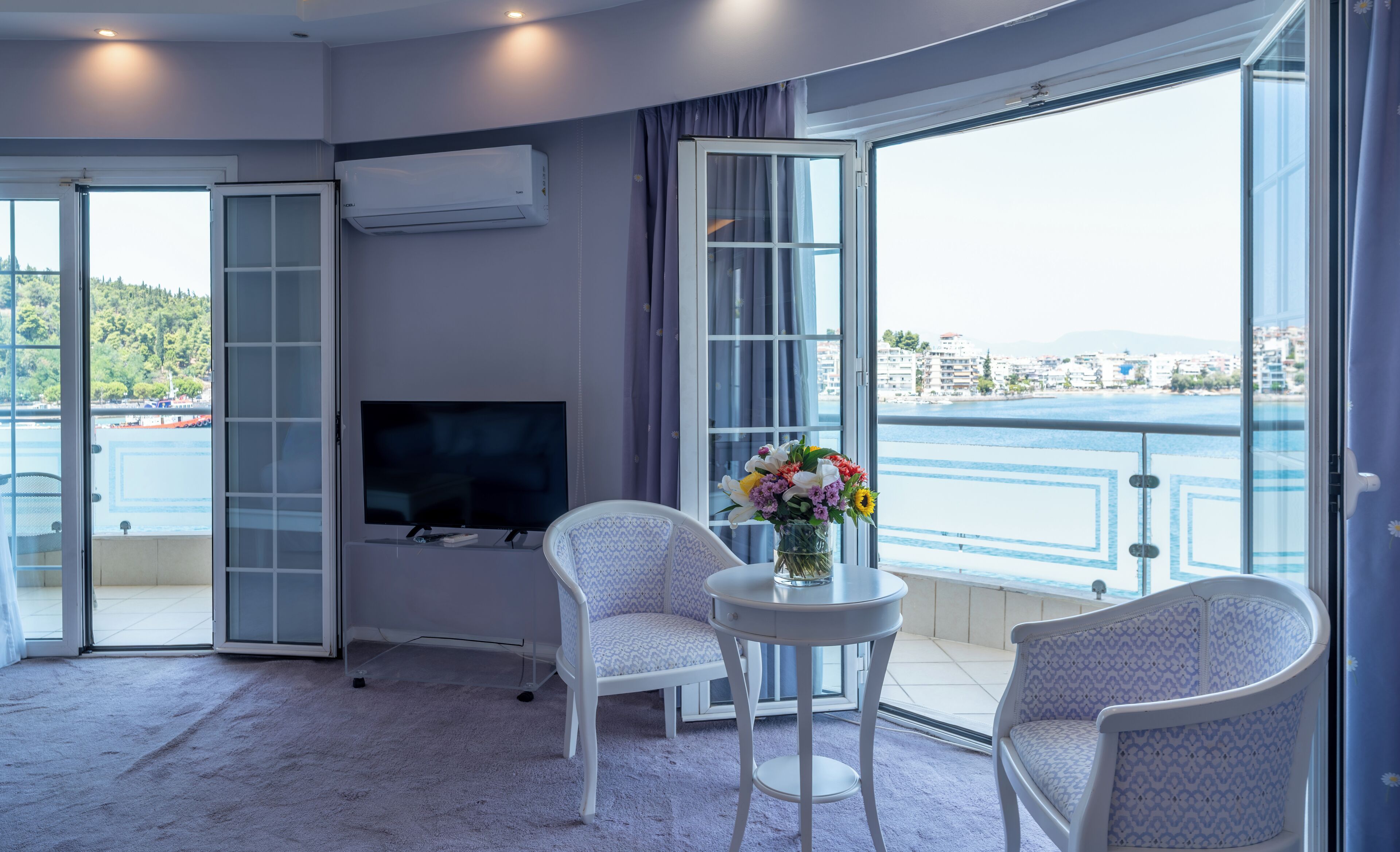 Quarto de hotel com televisão, duas cadeiras, uma mesa pequena com um vaso de flores e uma varanda com vista para o mar e edifícios.