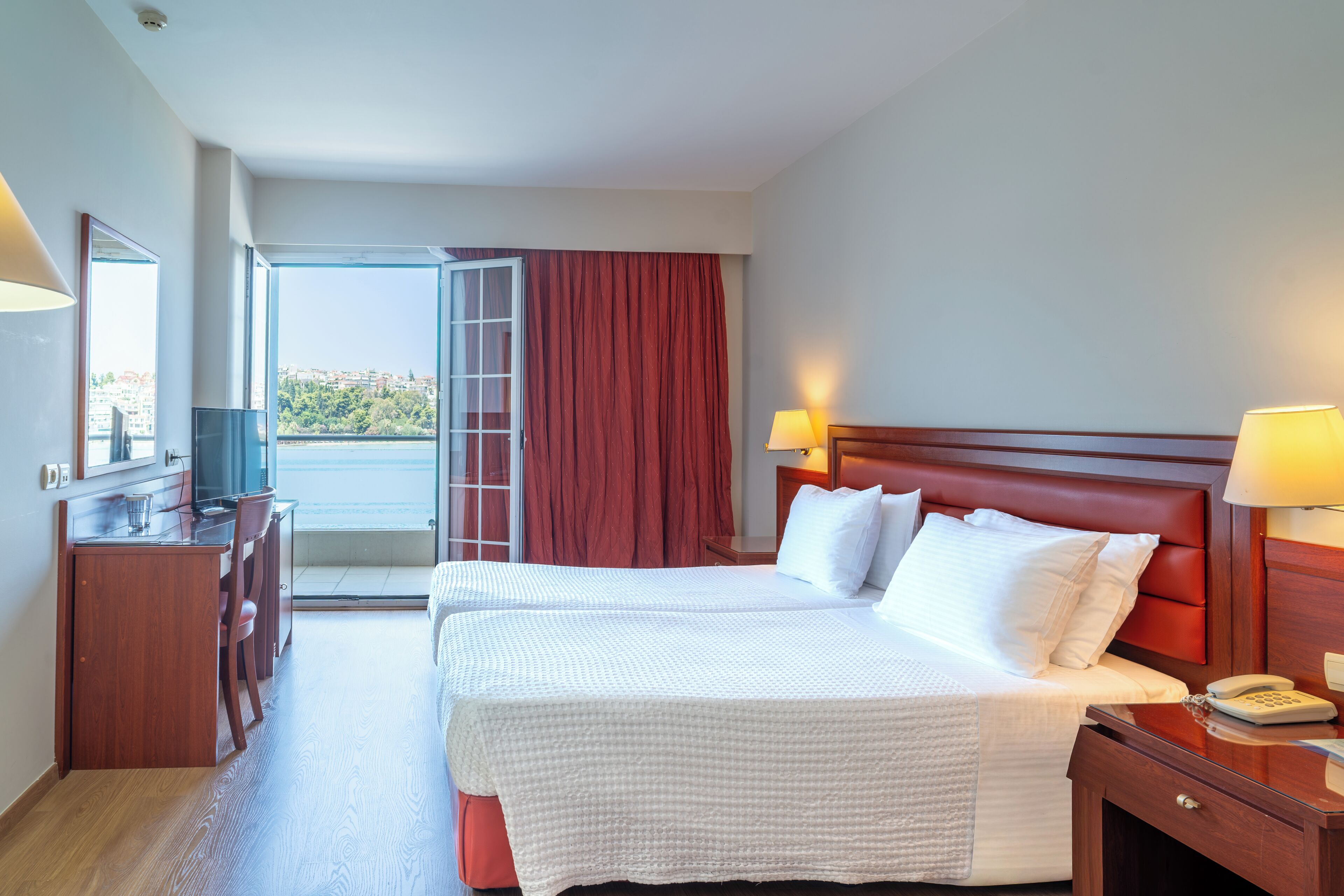 Quarto de hotel com cama, luminárias laterais, escrivaninha com telefone e vista para o exterior.