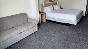Quarto de hotel com cama, sofá, mesa de cabeceira com luminária e uma obra de arte na parede.