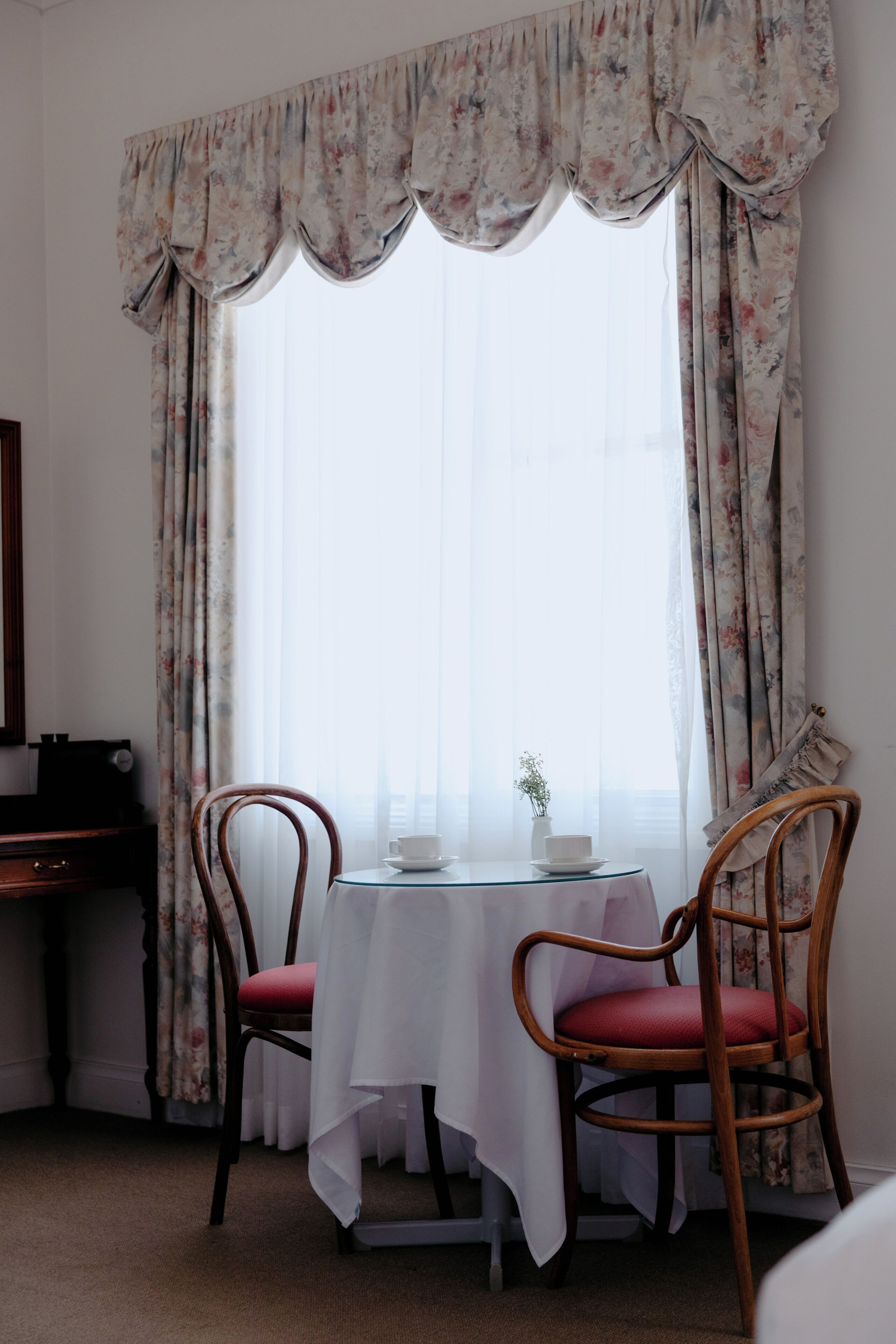 Quarto com uma mesa de jantar posta para dois, duas cadeiras de madeira com almofadas vermelhas, uma pequena planta em vaso sobre a mesa e uma janela com cortinas florais.