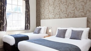 Quarto de hotel com duas camas, janela com cortinas, luminária e papel de parede estampado.