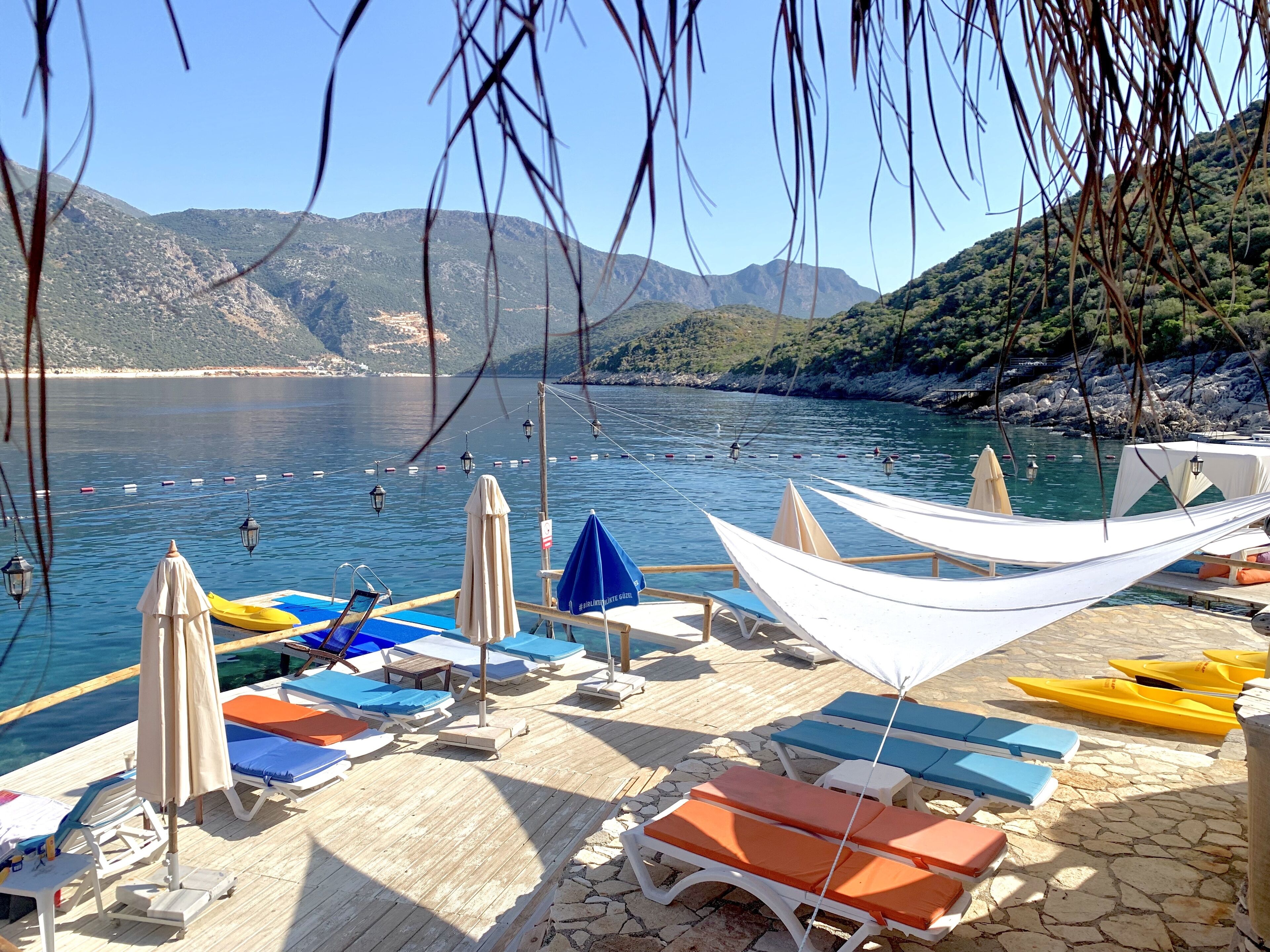 Uma praia com espreguiçadeiras e guarda-sóis, um deck de madeira e vista para o mar e montanhas.