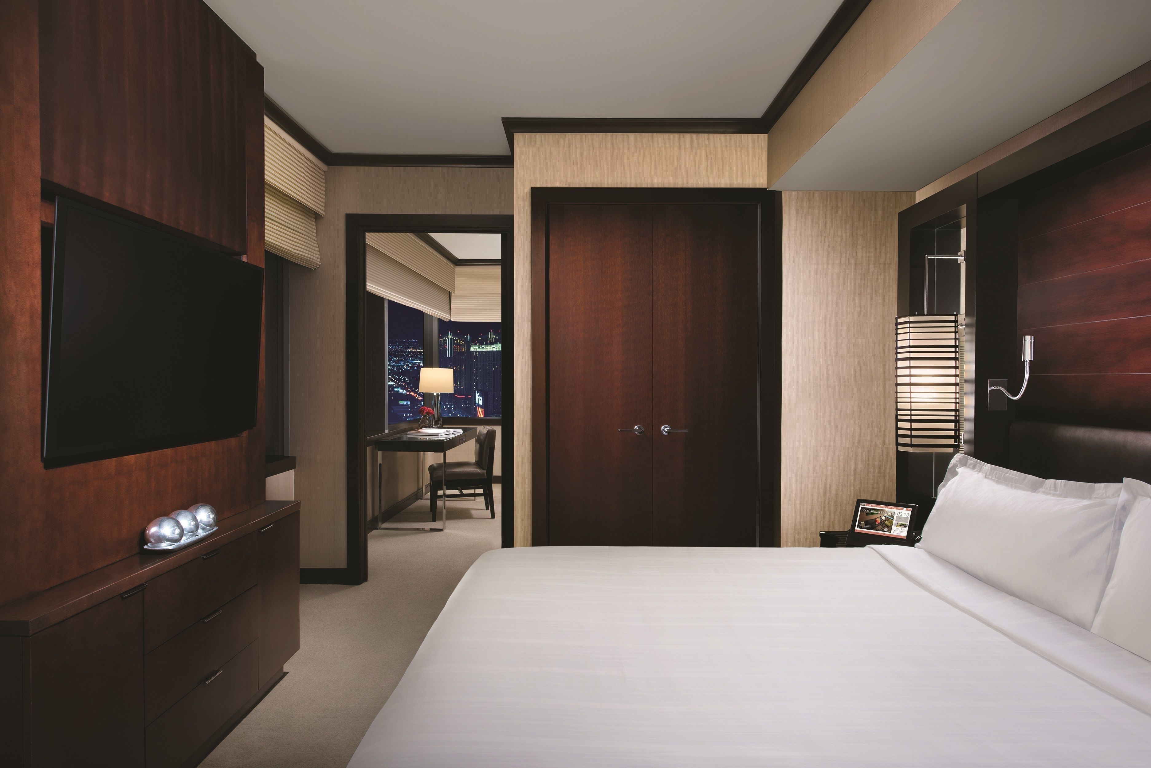 Quarto de hotel com uma cama grande, uma TV sobre uma cômoda de madeira, uma escrivaninha com uma cadeira e vista para a cidade à noite.