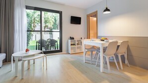 Uma sala de estar moderna com um sofá branco, uma pequena mesa redonda, um espaço de refeições com mesa e cadeiras brancas, uma TV fixada na parede e um grande painel de vidro deslizante que dá para uma varanda com mobiliário de exterior.