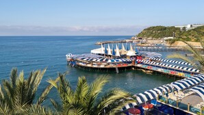 Uma marina com vários barcos atracados, um cais que se estende sobre a água e uma área de praia com edifícios ao fundo.