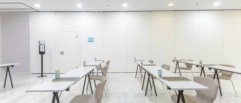 Uma sala de conferências com mesas brancas, cadeiras e um quadro branco.