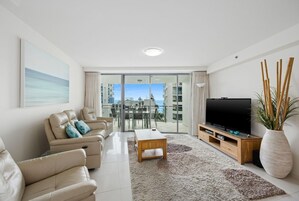 Uma sala de estar moderna com um conjunto de sofá, uma mesa de centro, uma TV e uma janela grande com vista para edifícios e um céu limpo.