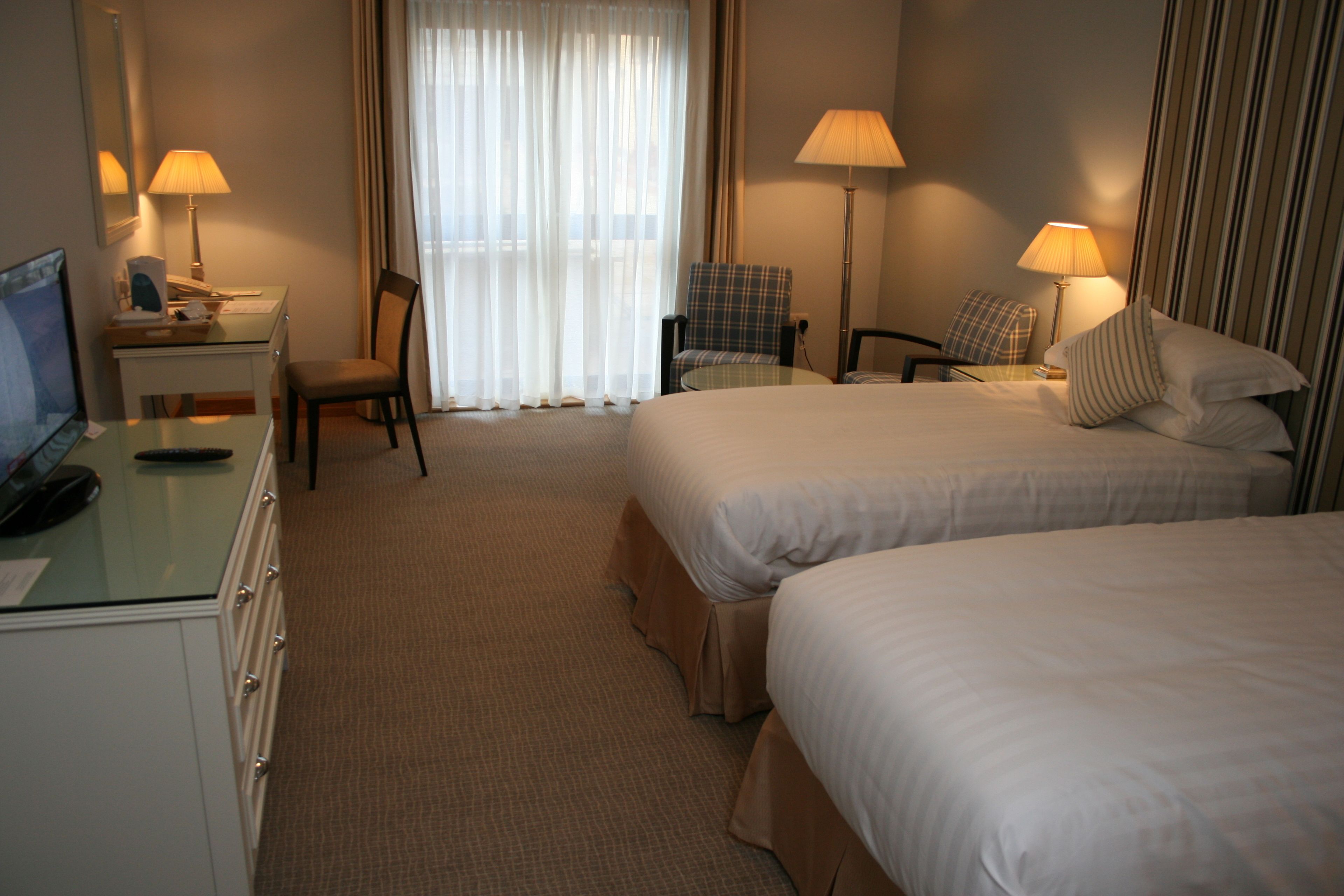 Quarto de hotel com duas camas, uma mesa com computador, uma cadeira e um abajur.