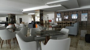 Espaço interior moderno com diversas áreas de estar, incluindo mesas e cadeiras, e uma parede de tijolos com cartazes informativos.