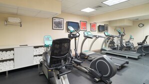 Academia com diversos equipamentos de exercício, incluindo esteiras e bicicletas ergométricas, e uma televisão fixada na parede.