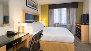 Quarto de hotel com cama, mesa de cabeceira com luminária, escrivaninha com telefone, cadeira, televisão e janela com cortinas.