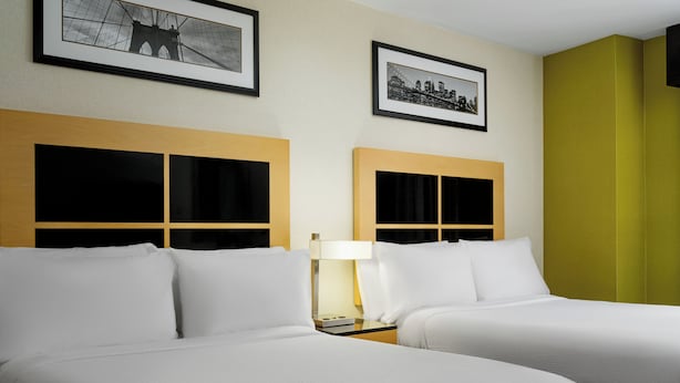 Quarto de hotel com duas camas, uma mesinha de cabeceira com abajur e duas imagens emolduradas na parede.
