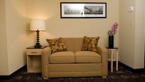Quarto de hotel com um sofá bege, uma mesinha com luminária, um quadro na parede e um vaso com flores.