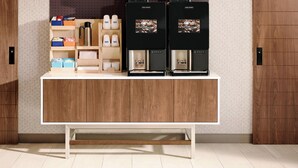 Uma estação de café com duas máquinas de café, uma prateleira com chávenas e um balcão com diversos artigos.