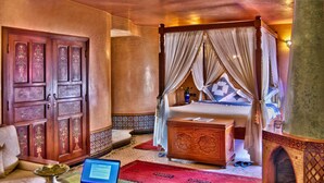 Quarto tradicional com cama de dossel, um armário de madeira, um laptop sobre uma pequena mesa e um tapete vermelho estampado.