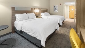 Quarto de hotel com duas camas, uma escrivaninha, uma cadeira e uma porta de madeira.