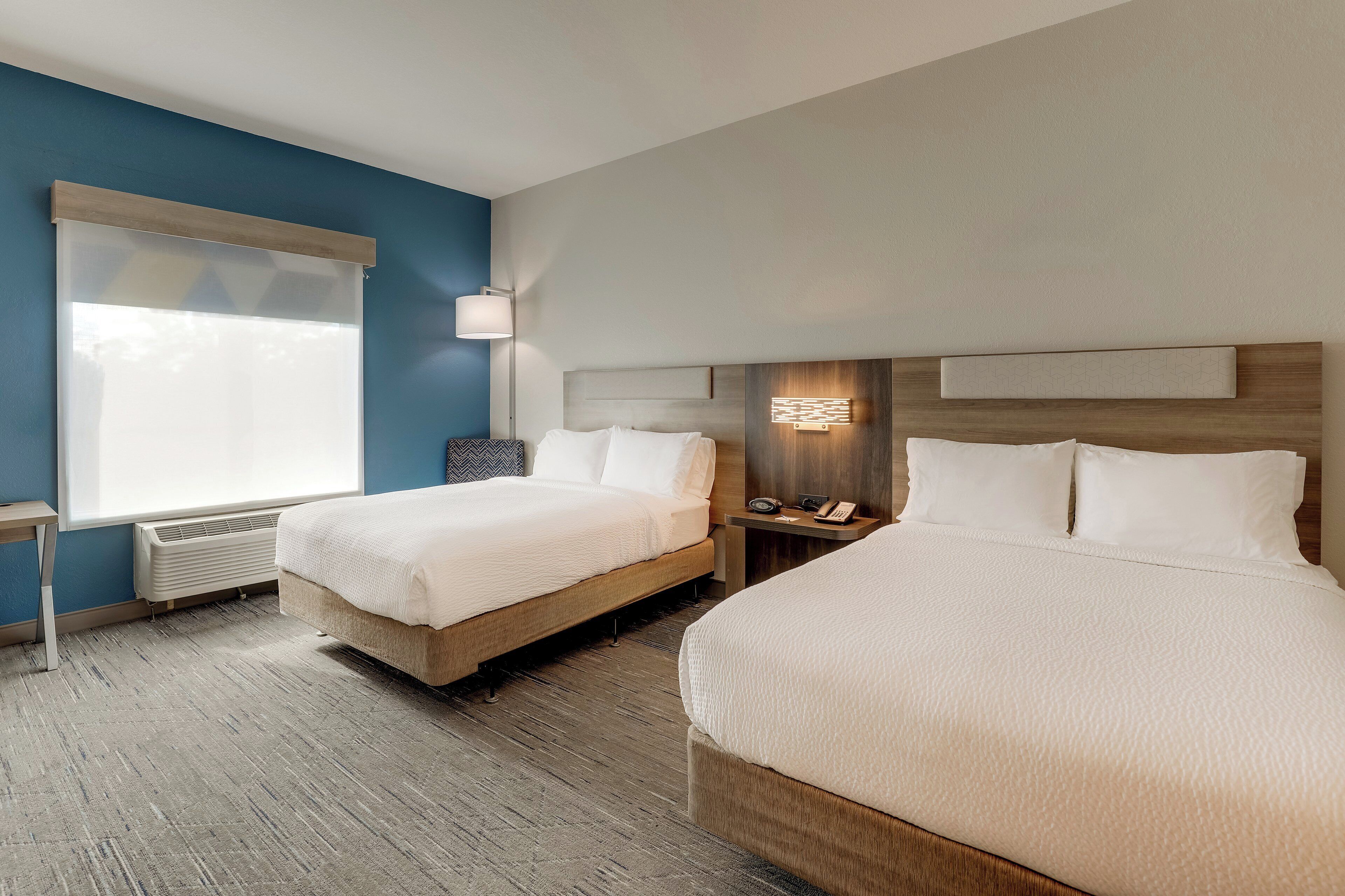 Quarto de hotel com duas camas, mesa de cabeceira, abajur e janela com persianas.