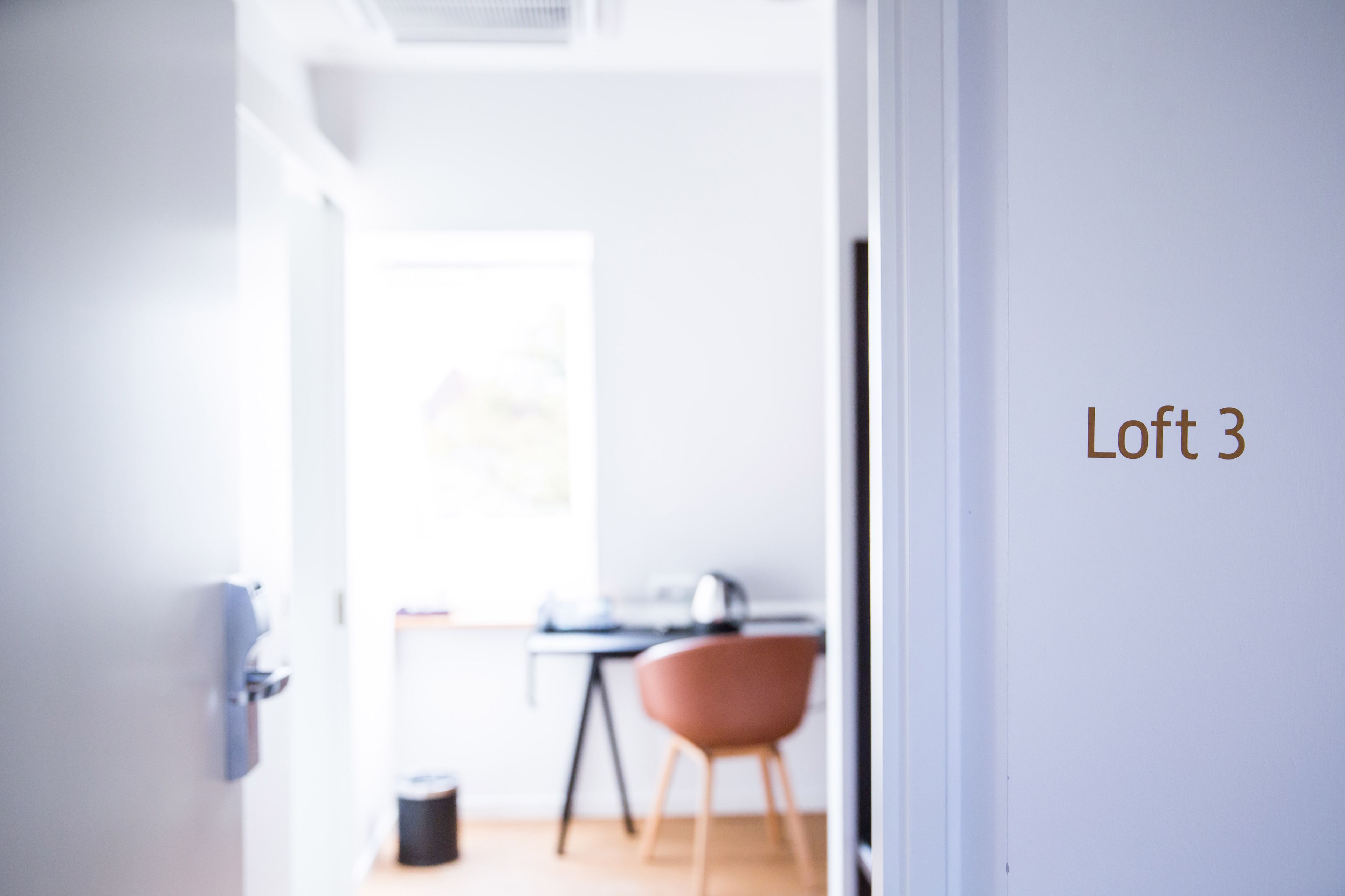 Vista interna do Loft 3, com uma porta, uma escrivaninha, uma cadeira e uma janela.