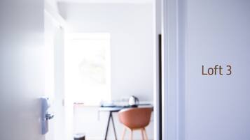 Vista interna do Loft 3, com uma porta, uma escrivaninha, uma cadeira e uma janela.