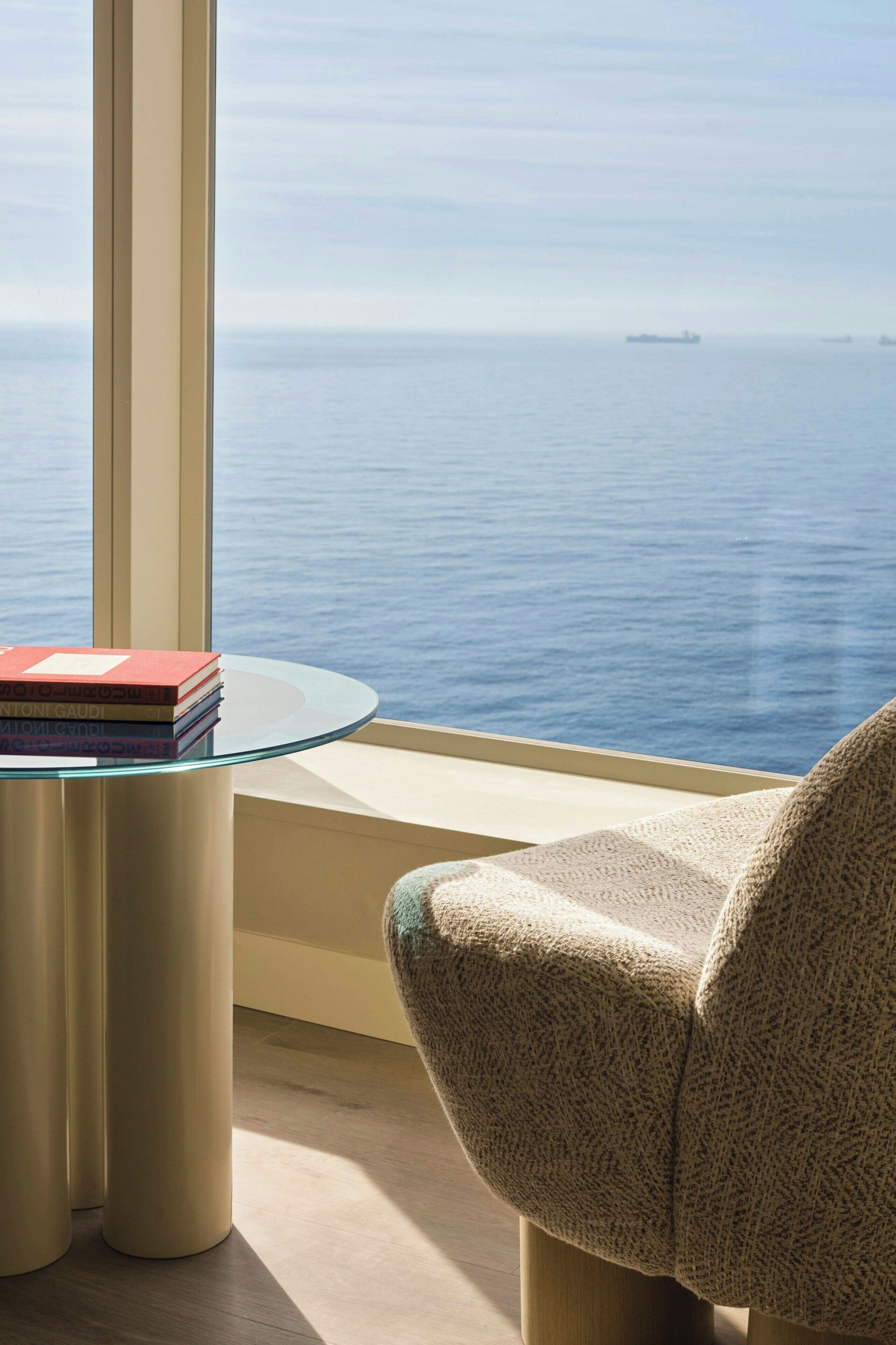 Quarto com uma janela grande, uma mesa de vidro redonda com livros, uma poltrona bege e vista para o mar.