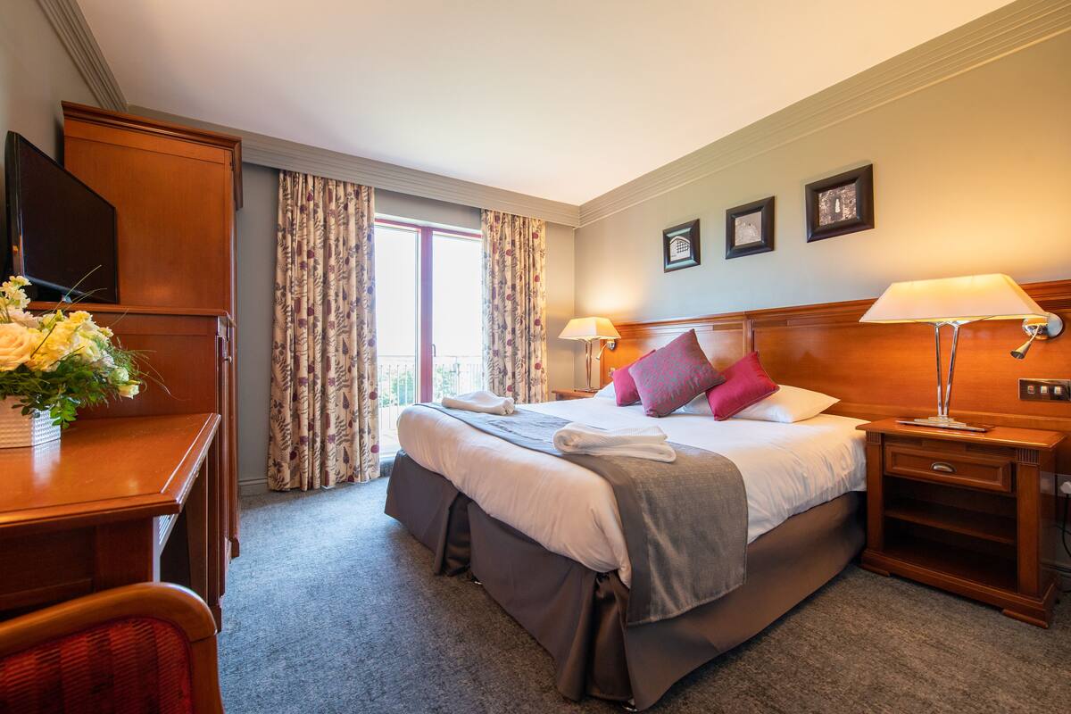 Quarto de hotel com uma cama grande, abajures, escrivaninha com televisão e janela com cortinas.