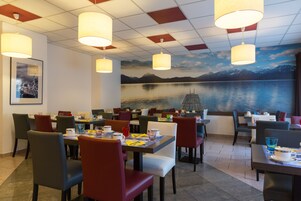 Um restaurante com várias mesas e cadeiras, um mural com um lago e montanhas, e luminárias pendentes.