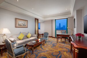 Quarto de hotel com sofá, poltronas, mesa de centro, escrivaninha e uma janela grande com vista para a cidade.