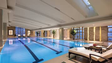 Uma grande piscina interior com água azul cristalina, rodeada por espreguiçadeiras e uma área de estar com uma mesa.