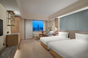 Quarto de hotel com duas camas, uma escrivaninha, uma cadeira e uma janela grande com vista para a cidade.