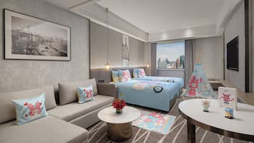 Quarto de hotel moderno com vista para a cidade, uma cama grande com roupa de cama temática de desenhos animados, um sofá com almofadas combinando, uma mesa de centro redonda e uma pequena mesa com figuras.