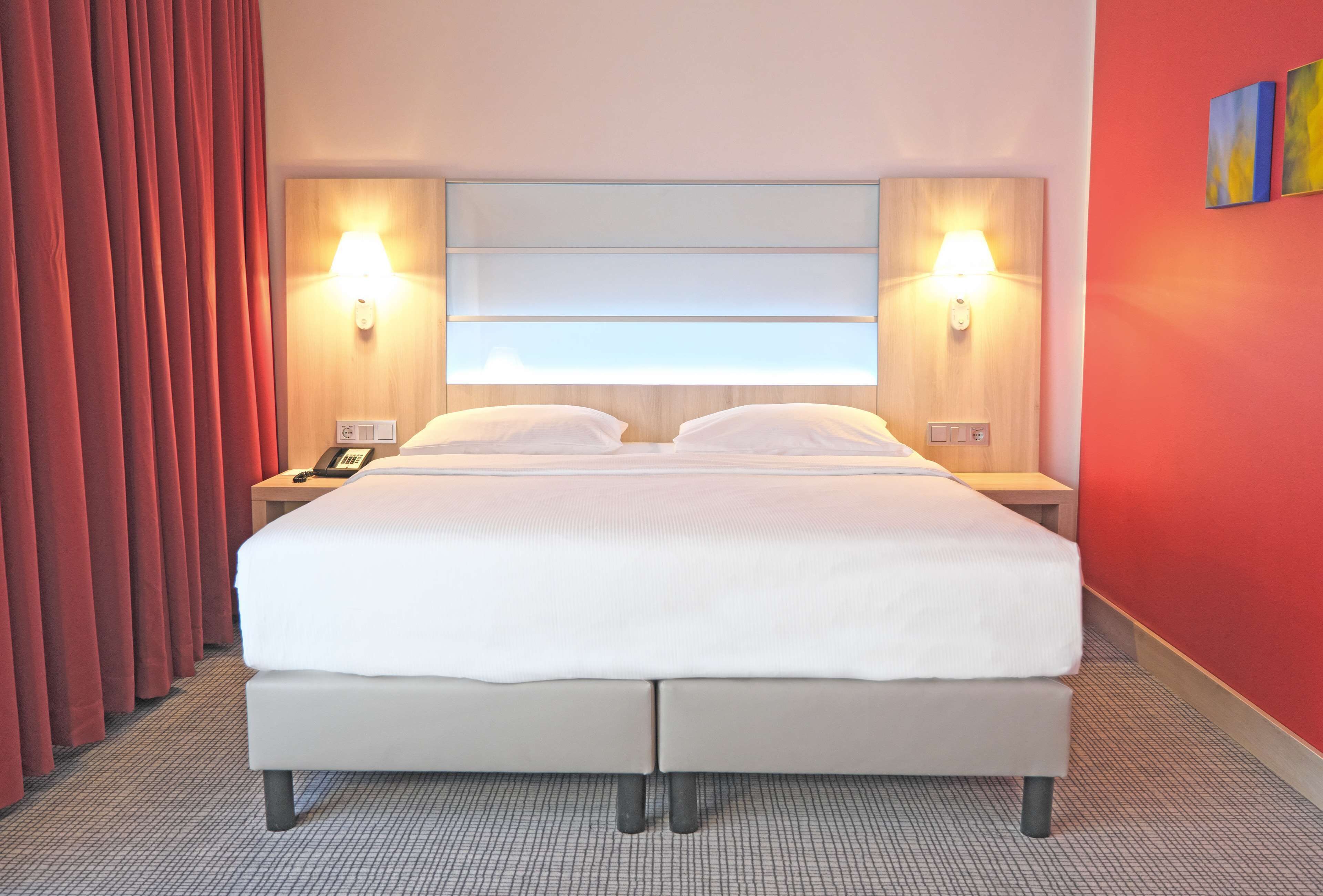 Uma cama de casal com cabeceira, duas mesas de cabeceira com abajures, um telefone e uma cortina vermelha.