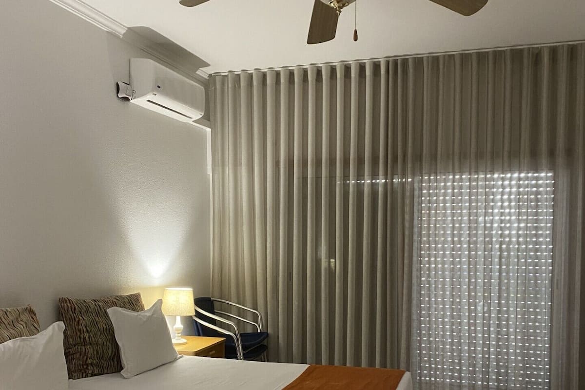 Quarto de hotel com cama, ventoinha no teto, janela com persianas e mesa de cabeceira com abajur.