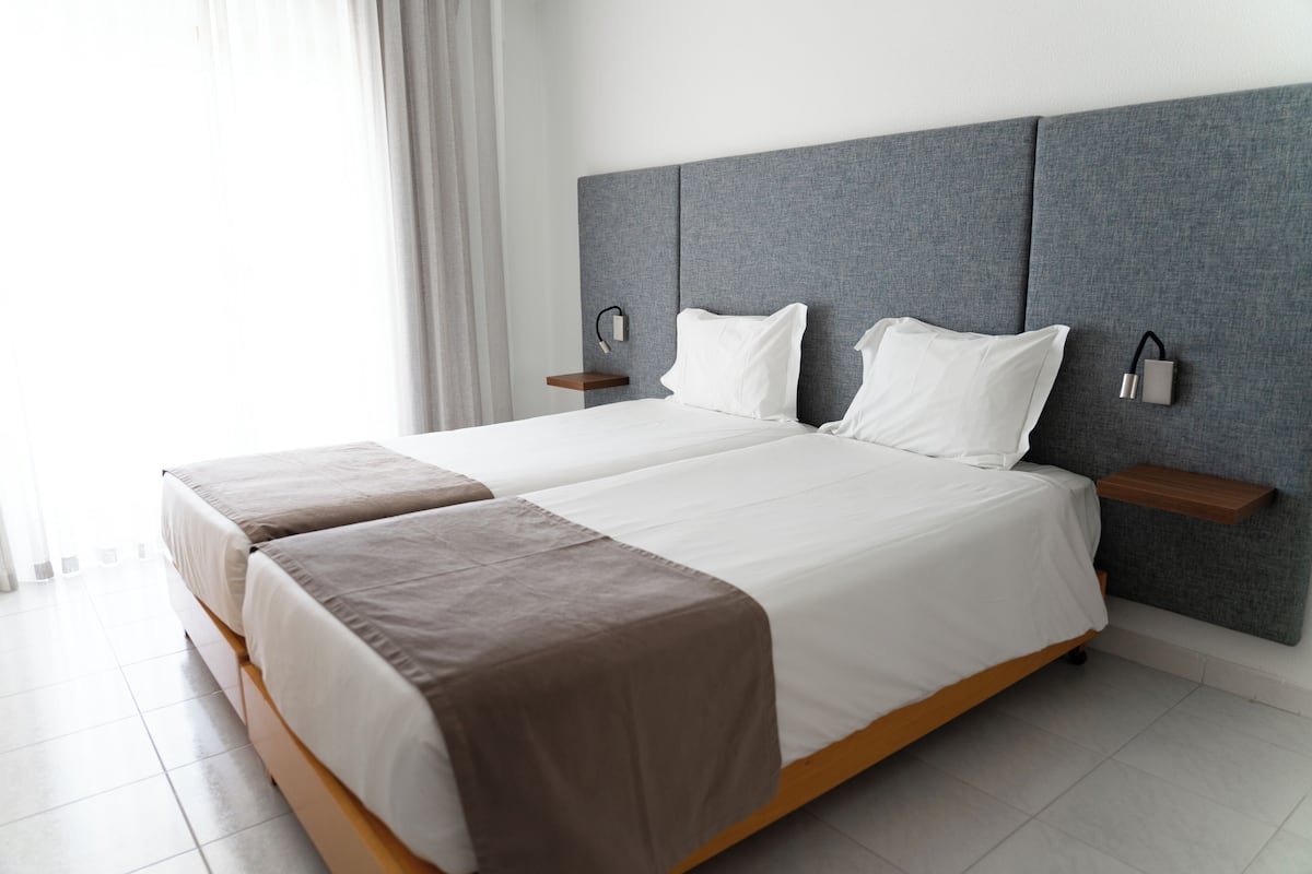 Cama de casal com cabeceira cinza, roupa de cama branca e mesas de cabeceira com abajures.