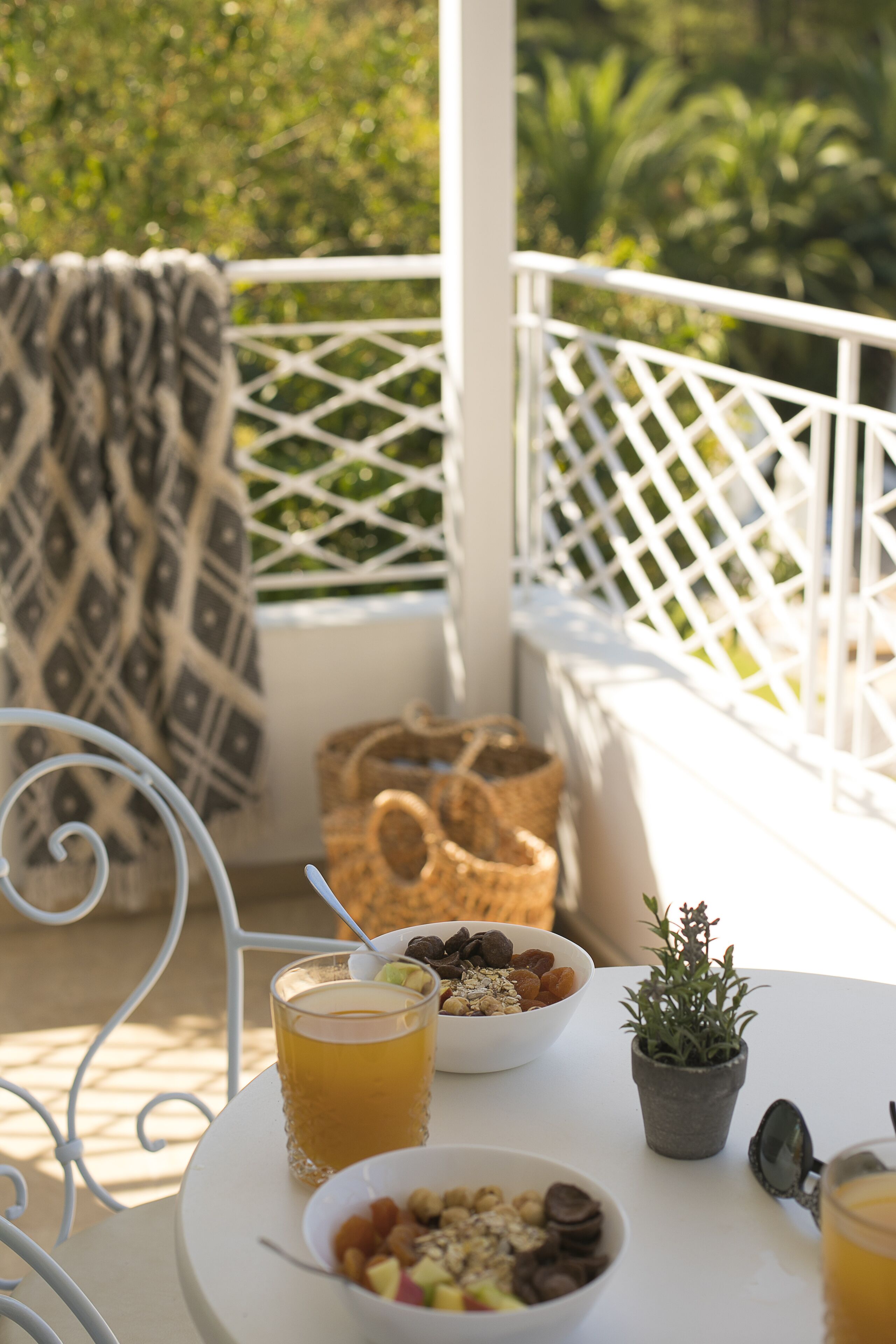 Uma mesa com uma bebida, taças de frutos secos e fruta desidratada, uma pequena planta em vaso e um par de óculos de sol numa varanda com guarda de proteção branca e um cobertor estampado.