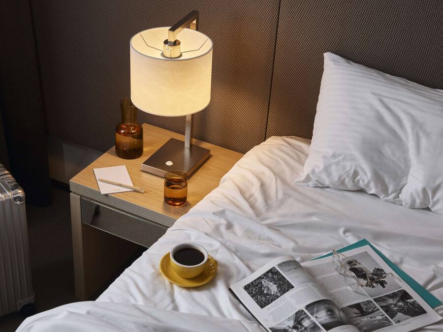 Cama bem arrumada com lençóis brancos, abajur, xícara de café e uma revista sobre a cama.