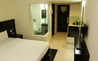 Quarto de hotel com cama, banheiro com box de vidro, escrivaninha com computador e televisão.