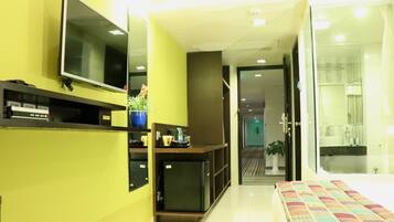 Quarto de hotel com cama, televisão fixada na parede, minibar e prateleira com planta, além de um banheiro visível através de uma porta aberta.