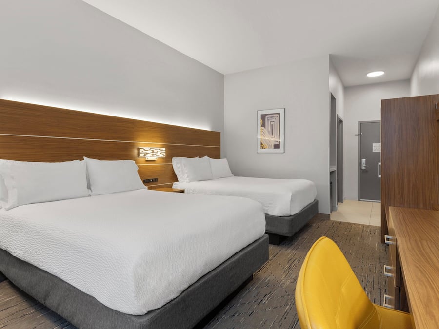 Quarto de hotel moderno com duas camas, cabeceira de madeira, uma cadeira amarela e televisão de tela plana.