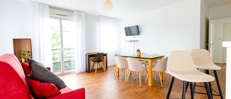 Uma sala de estar moderna com um sofá vermelho, uma mesa de jantar com cadeiras, uma TV fixada na parede e uma varanda visível pela janela.