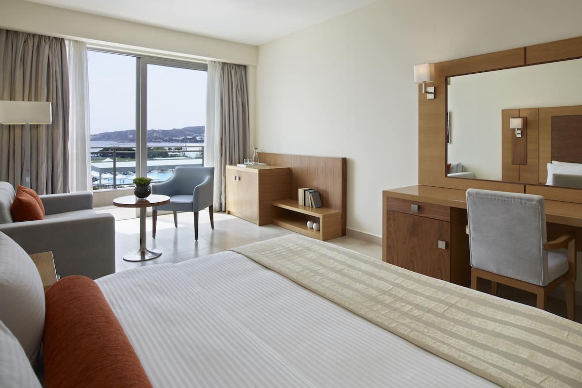 Quarto de hotel com uma cama grande, uma escrivaninha, uma cadeira, uma mesinha e vista da cidade através de grandes janelas.