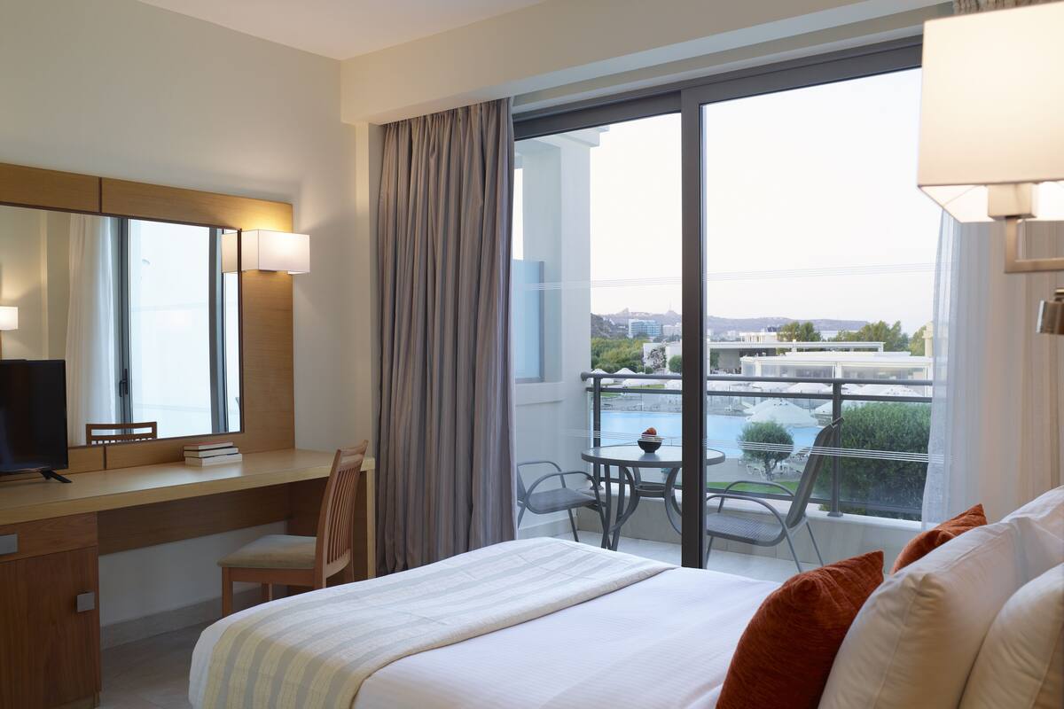 Quarto de hotel com cama, escrivaninha, cadeira, televisão e varanda com vista para a cidade.