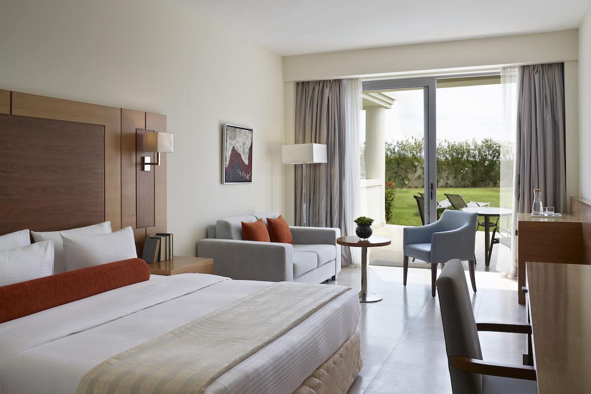 Quarto de hotel com uma cama grande, um sofá, uma mesa pequena e uma cadeira. Tem uma varanda com vista para o jardim.