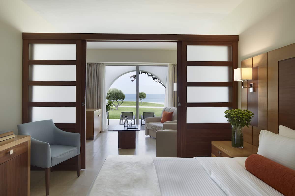 Quarto de hotel com cama, escrivaninha, cadeira, sofá e vista para o mar através de uma porta aberta.