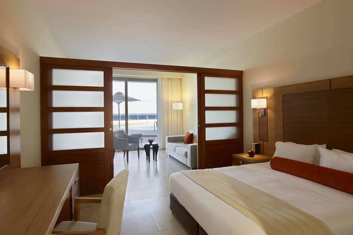 Quarto de hotel com cama, escrivaninha, cadeira, mesa pequena e vista para o mar através de uma porta de vidro.