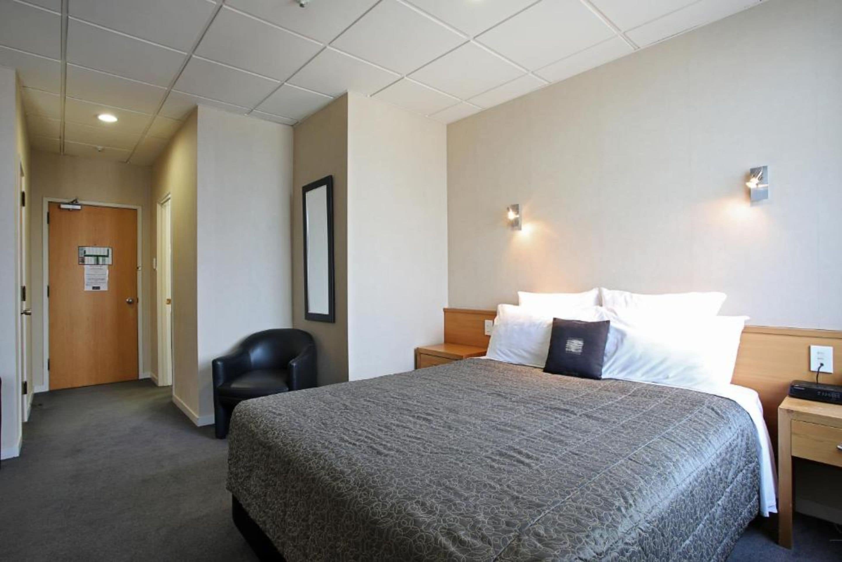 Quarto de hotel com uma cama grande, uma mesa de cabeceira, uma cadeira e uma porta que dá para outro espaço.